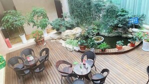 Uma área de pátio ao ar livre com piso de madeira, cadeiras de vime e mesas. Há um pequeno lago rodeado por pedras e diversos vasos de plantas.