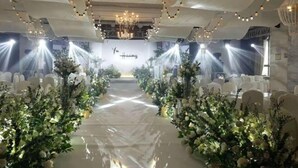 Espaço para eventos formais com uma passagem central ladeada por arranjos florais, mesas montadas para convidados e lustres.