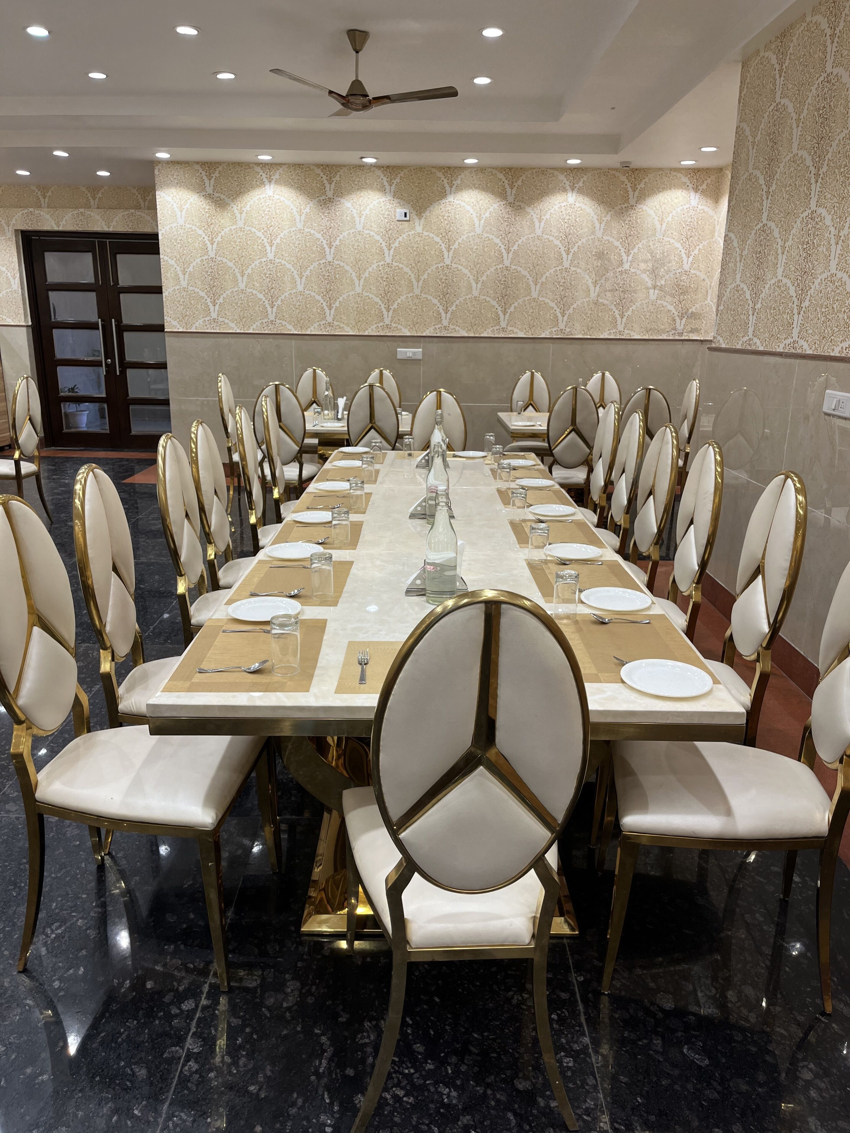 Uma sala de conferência com uma mesa retangular comprida, cadeiras brancas com detalhes dourados e um ventilador de teto.