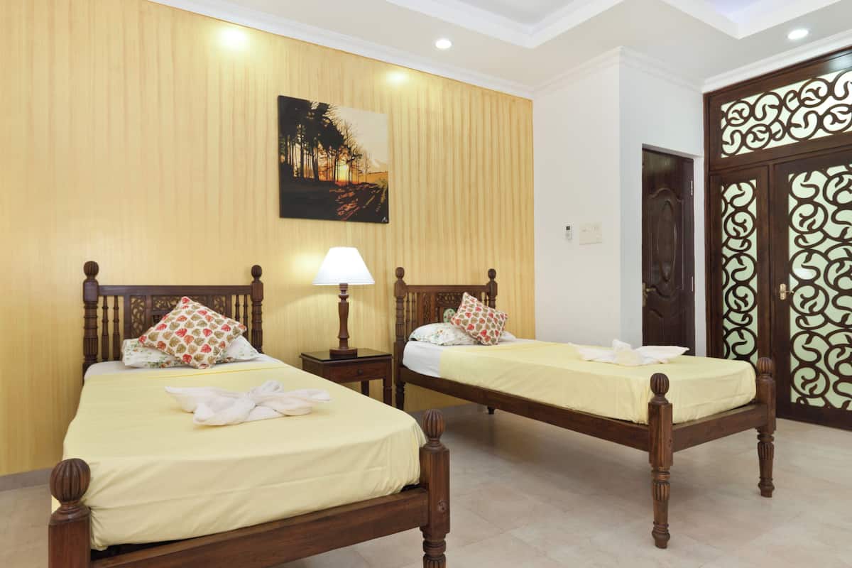 Quarto de hotel com duas camas de madeira, cada uma com colcha e almofadas amarelas, uma mesa de cabeceira com abajur e um quadro na parede.
