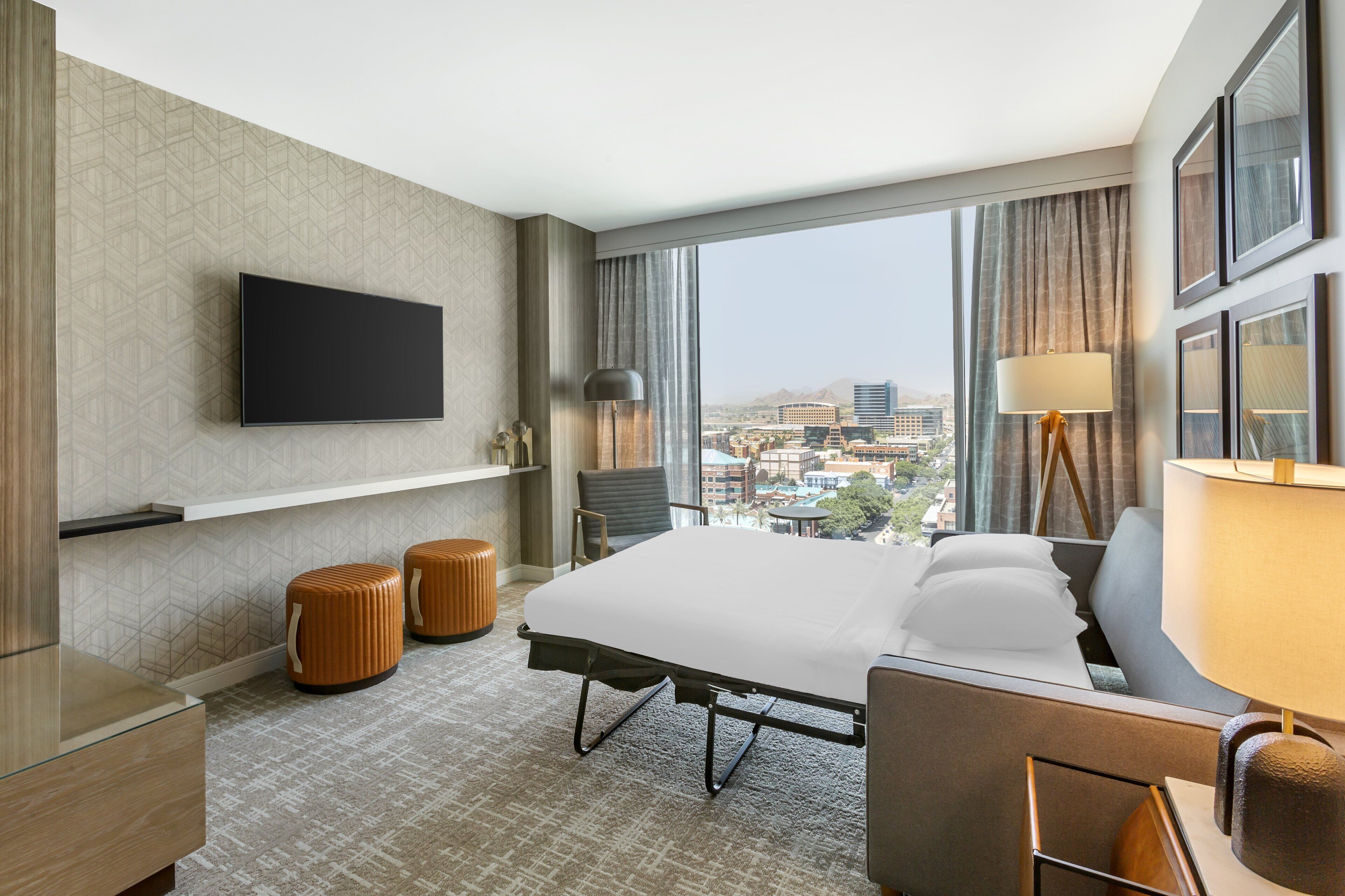Quarto de hotel com uma cama grande, uma TV fixada na parede, uma escrivaninha com uma luminária e vista da cidade através de amplas janelas.