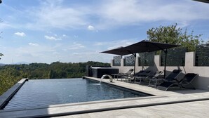 Uma piscina infinita retangular com cascata, rodeada por espreguiçadeiras e uma área de estar coberta.
