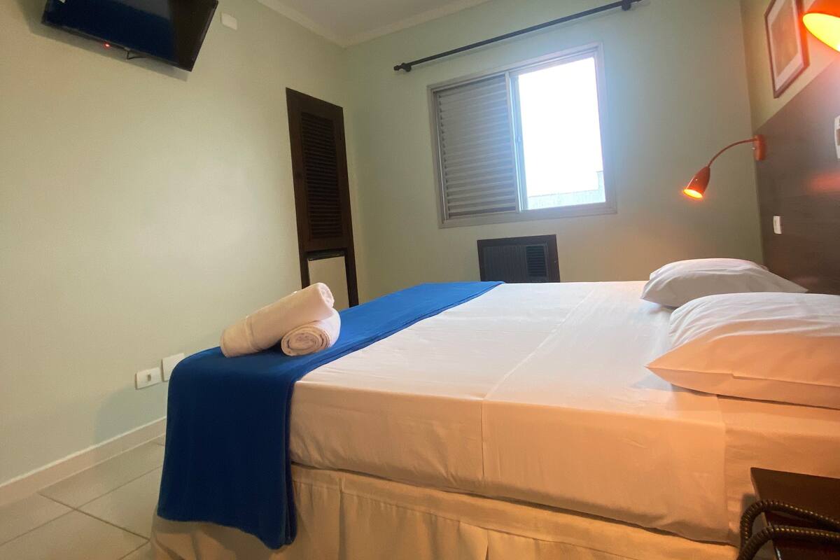Quarto de hotel com uma cama, televisão fixada na parede, janela com persianas e abajur.