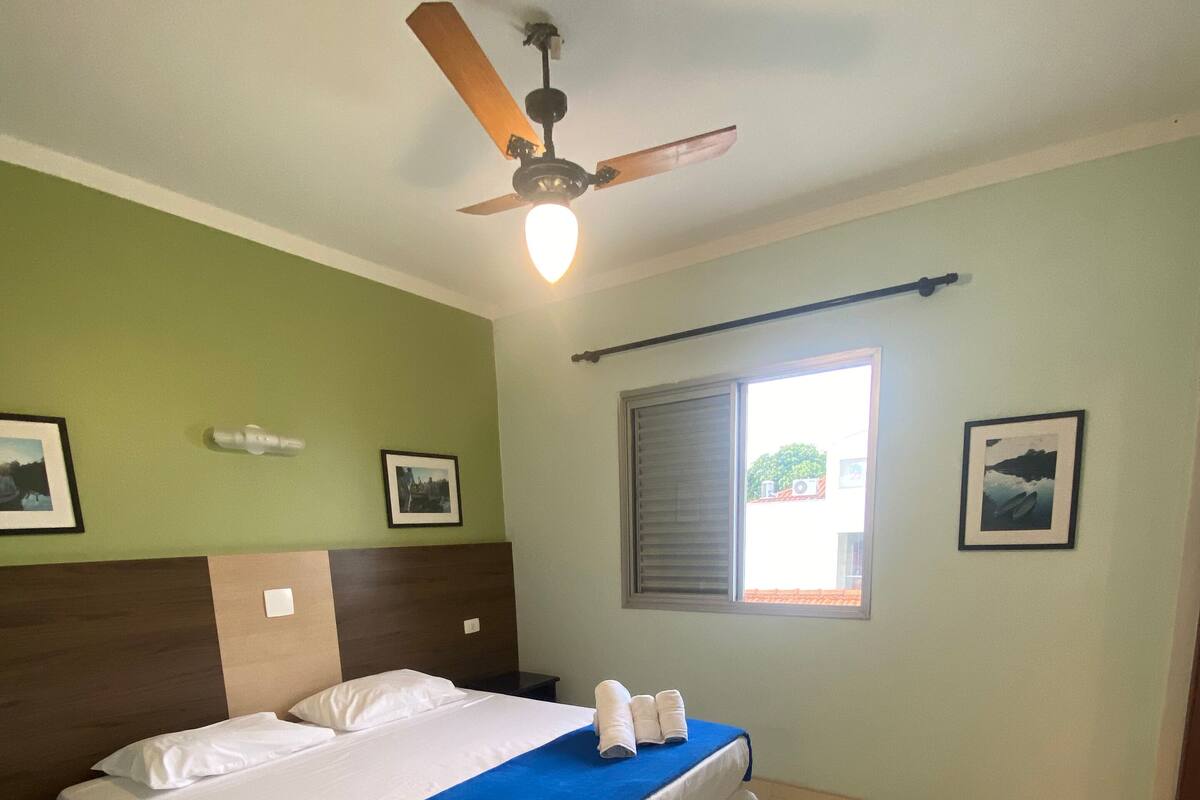 Quarto de hotel com cama, ventilador de teto, janela com persianas e quadros na parede.