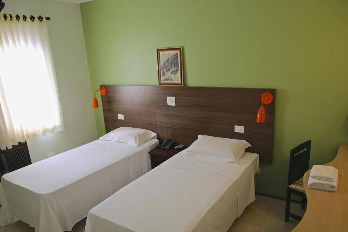 Quarto de hotel com duas camas de solteiro, cabeceira de madeira, um quadro, um telefone e uma cadeira.