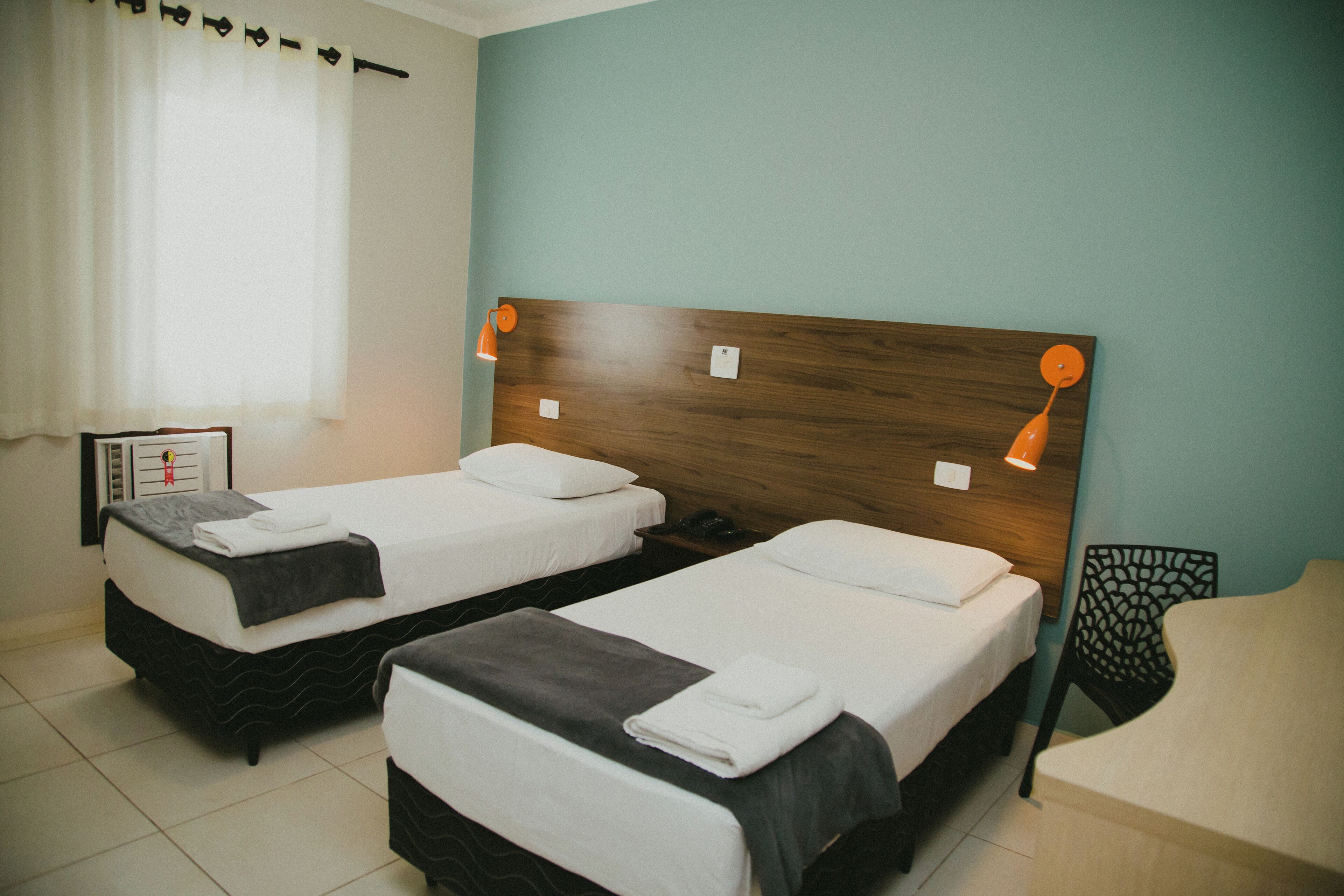 Quarto de hotel com duas camas de solteiro, cabeceira de madeira, mesa de cabeceira, uma cadeira e janela com cortinas.