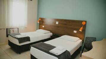 Quarto de hotel com duas camas de solteiro, cabeceira de madeira, mesa de cabeceira, uma cadeira e janela com cortinas.