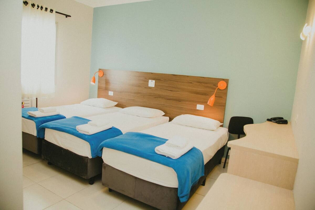 Quarto de hotel com três camas, cada uma com cobertor azul e lençóis brancos. Possui cabeceira de madeira, uma escrivaninha com cadeira e uma mesinha.