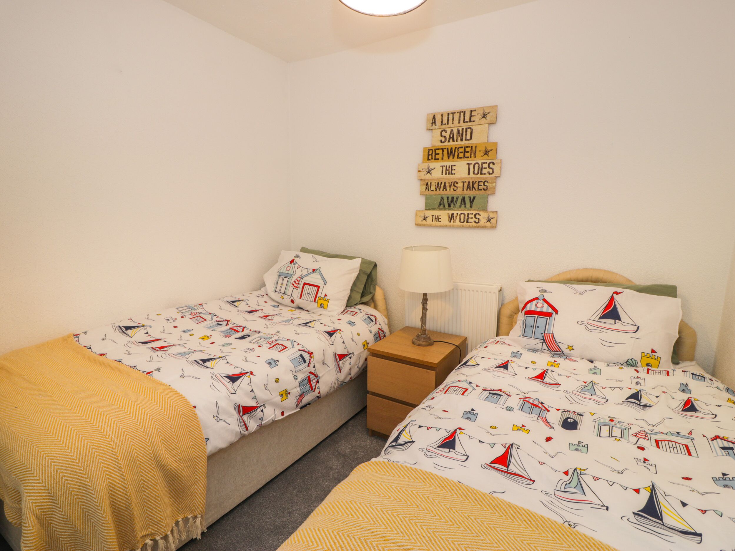 Quarto com duas camas de solteiro, cada uma com colcha de tema náutico, mesa de cabeceira com luminária e placa na parede com mensagem náutica.