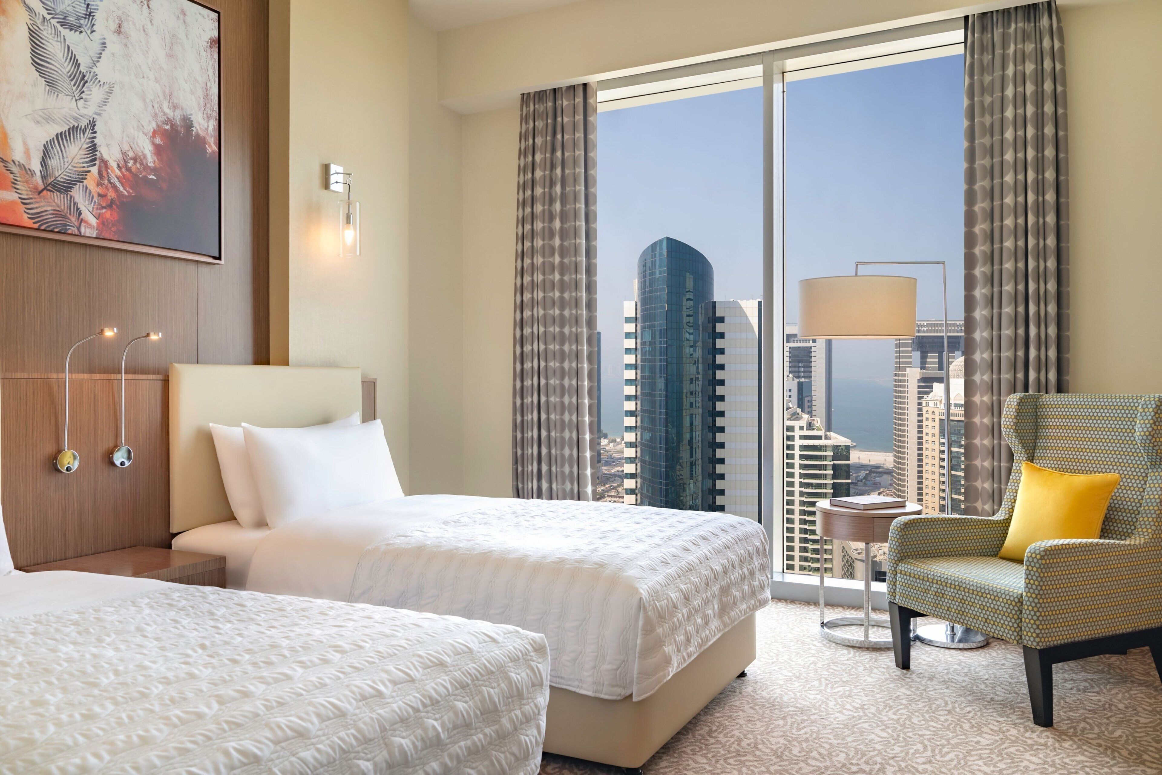 Quarto de hotel com duas camas, uma cadeira, um abajur e vista para a cidade.