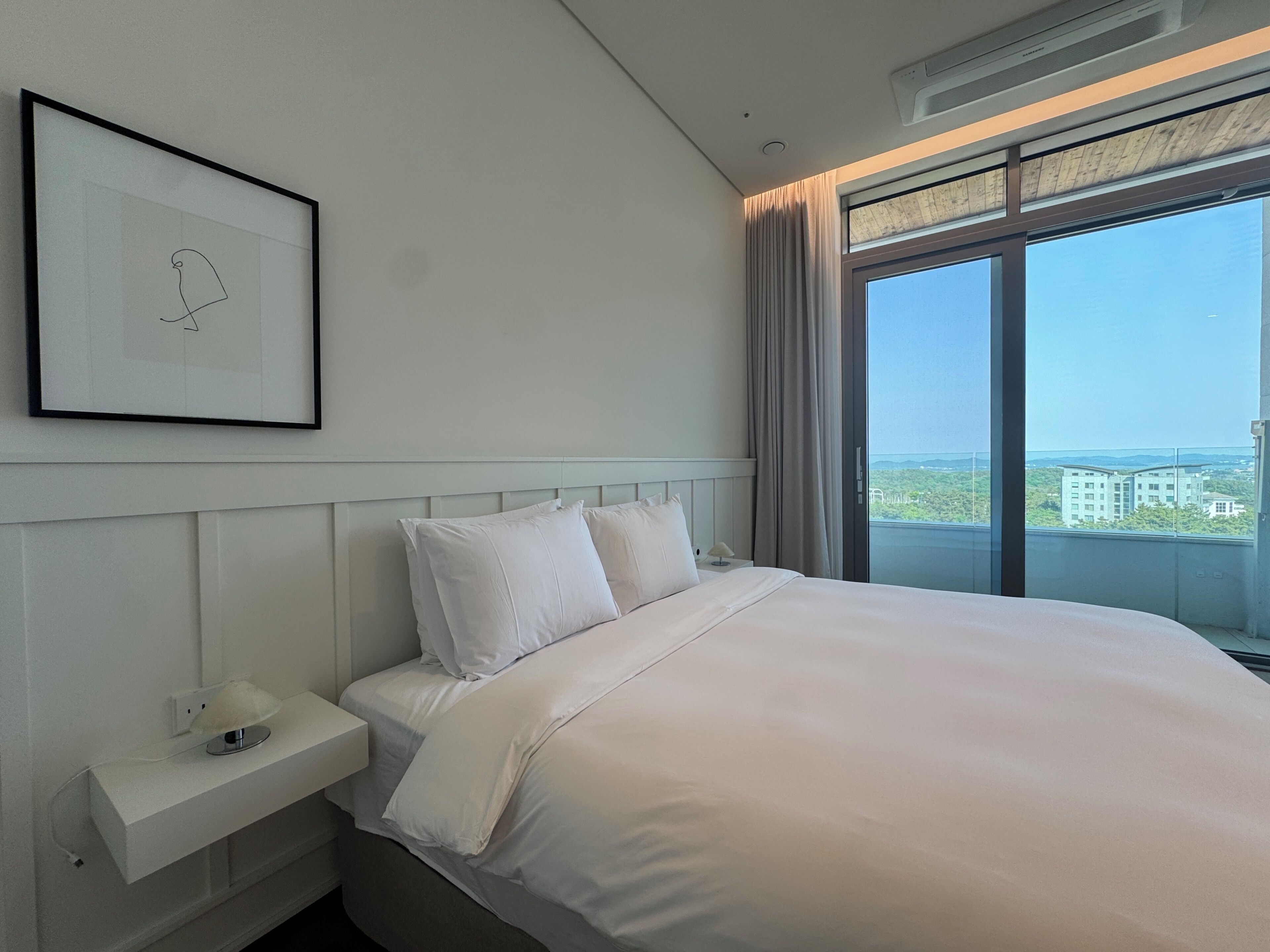 Quarto de hotel com cama, mesa de cabeceira, abajur, quadro emoldurado e uma grande janela com vista.