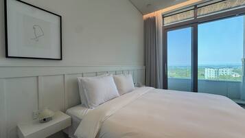Quarto de hotel com cama, mesa de cabeceira, abajur, quadro emoldurado e uma grande janela com vista.