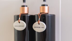 Duas embalagens pretas com bombas dosadoras, uma identificada como 'SHAMPOO' e a outra como 'SHOWER GEL', apoiadas sobre uma superfície branca, com um fundo de azulejos.