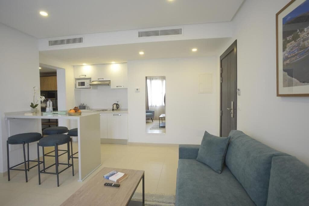 Uma sala de estar moderna com um sofá cinzento, uma pequena mesa de centro e uma área de cozinha com micro-ondas e chaleira.