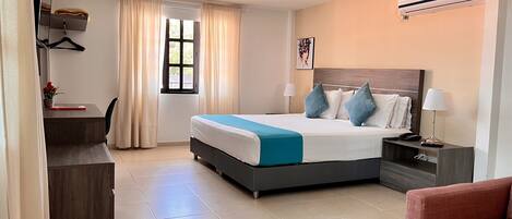 Quarto de hotel com cama, mesas de cabeceira, escrivaninha, cadeira, janela com cortinas e ar condicionado.