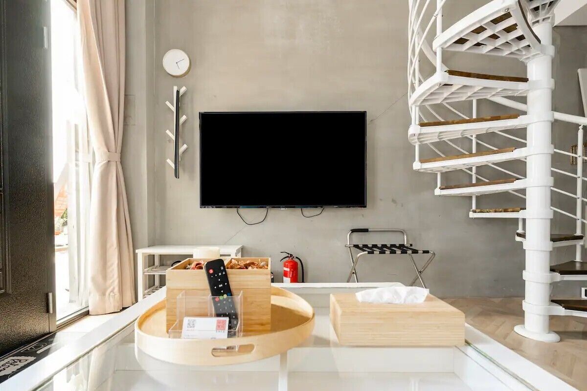 Uma sala de estar moderna com uma TV de tela plana na parede, uma mesinha de madeira com controles remotos, um extintor de incêndio e uma escada em espiral branca.