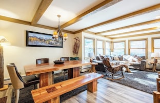 Uma sala de estar acolhedora com uma mesa de jantar de madeira, assentos confortáveis e grandes janelas com vista para o exterior.