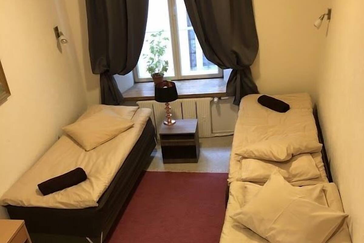 Quarto pequeno e compacto com duas camas simples, uma mesinha, uma cadeira e janela com cortinas.