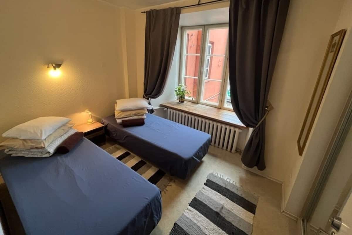 Quarto pequeno e limpo, com duas camas, janela com cortinas, radiador e luminária na parede.