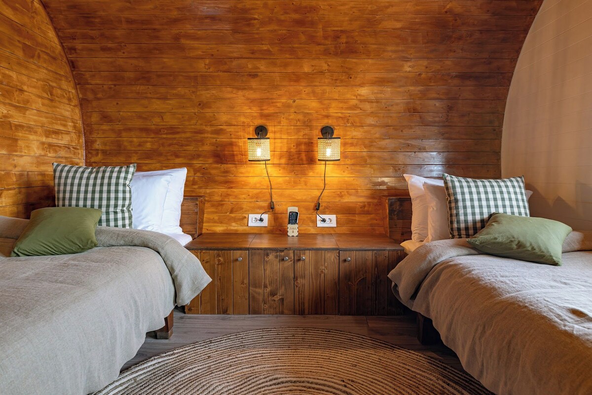 Um quarto acolhedor com paredes de madeira, duas camas com almofadas xadrez e luminárias fixadas na parede.