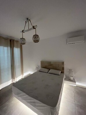 Quarto com cama, duas luminárias pendentes, ar condicionado fixado na parede e janela com cortinas.