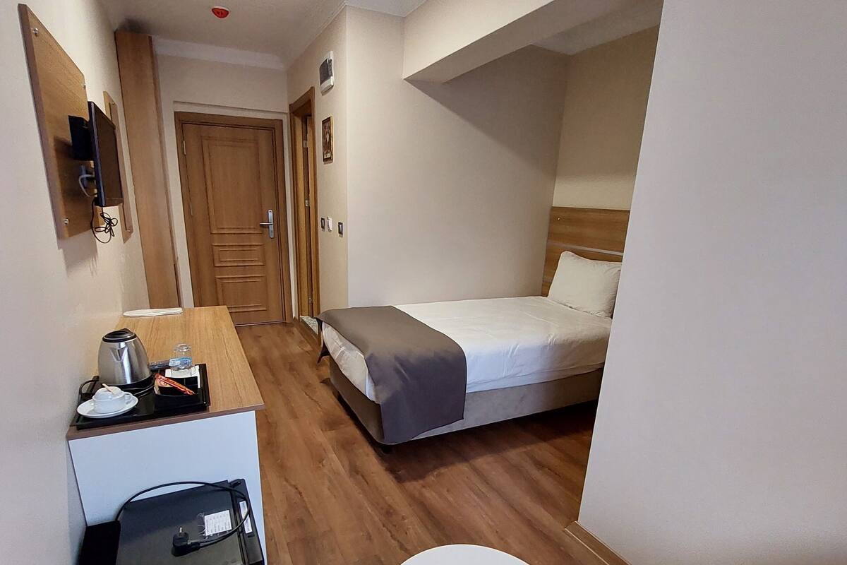 Quarto de hotel pequeno com uma cama, uma escrivaninha com chaleira, uma mesa branca e uma cadeira verde.