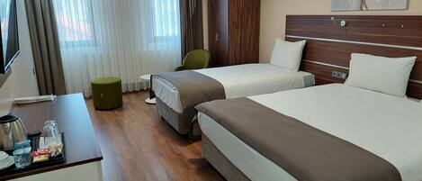 Quarto de hotel com duas camas, uma secretária com chaleira e chávenas, um banquinho verde e um quadro na parede.