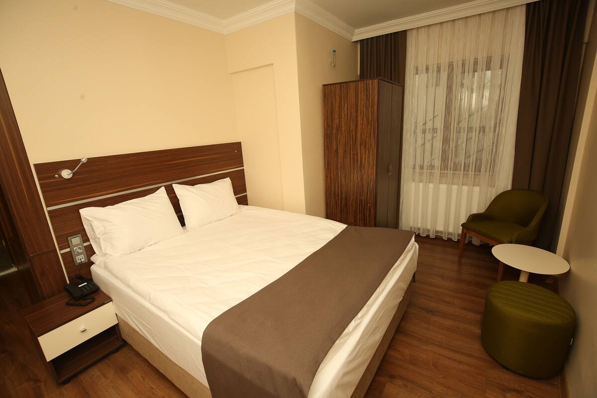 Quarto de hotel com uma cama grande, uma mesinha de cabeceira com telefone, um banquinho verde, uma pequena mesa redonda e uma cadeira.