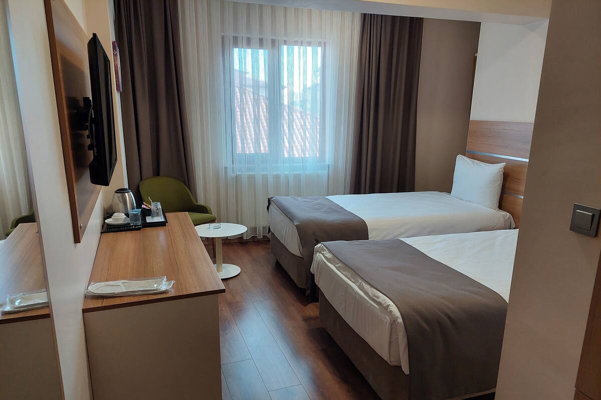 Quarto de hotel com duas camas, uma mesa redonda pequena, uma cadeira, cafeteira e televisão na parede.