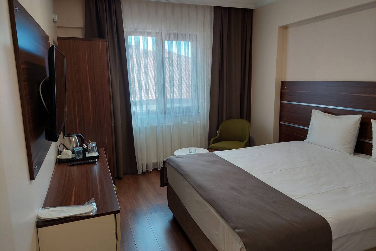 Quarto de hotel com uma cama grande, uma escrivaninha com televisão, uma cadeira e uma janela com cortinas.