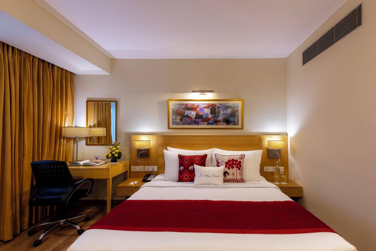 Quarto de hotel com cama, escrivaninha, cadeira, mesa de cabeceira, luminária, um quadro e janela com cortinas.