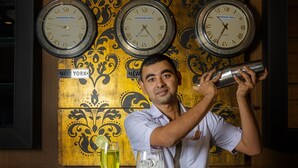 Um bar com um bartender preparando uma bebida, três relógios na parede e uma placa com a inscrição 'New York'.