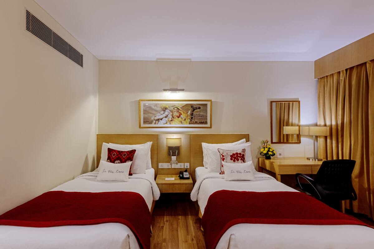Quarto de hotel com duas camas, uma escrivaninha, uma cadeira, um abajur, um quadro e um espelho.