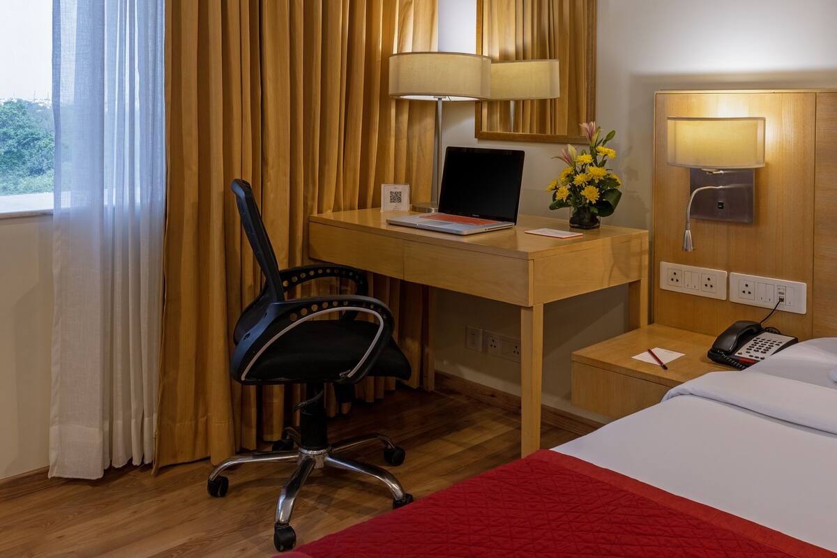 Quarto de hotel com cama, escrivaninha, cadeira, computador, abajur e telefone.