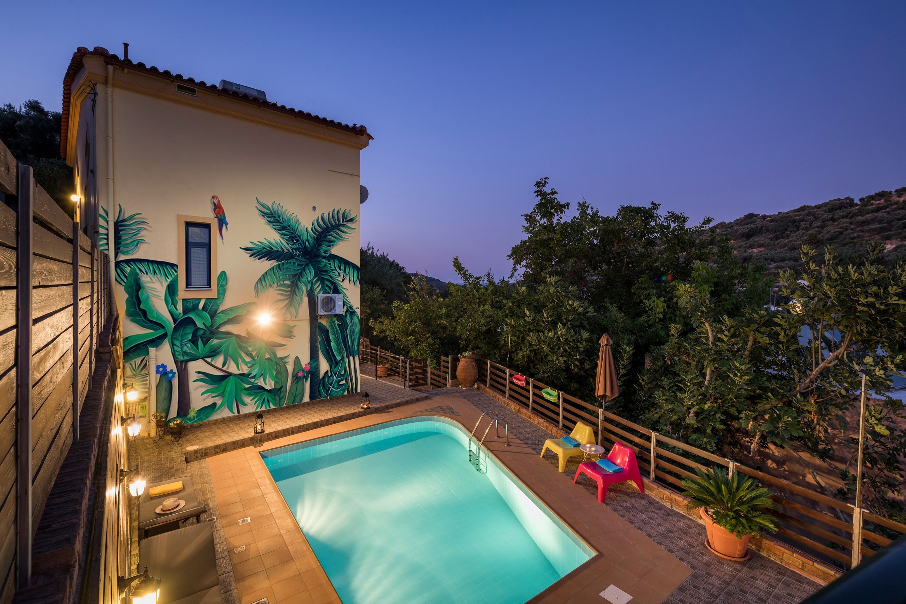Piscina com um mural de folhas tropicais e um papagaio, rodeada por cadeiras coloridas e vasos de plantas.