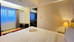 Quarto de hotel com uma cama grande, uma mesa, uma cadeira, uma luminária e vista para o exterior pela janela.