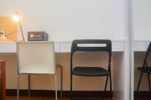 Quarto com uma cadeira branca, uma cadeira dobrável preta, uma escrivaninha com luminária e uma caixa com a etiqueta "Travelio".