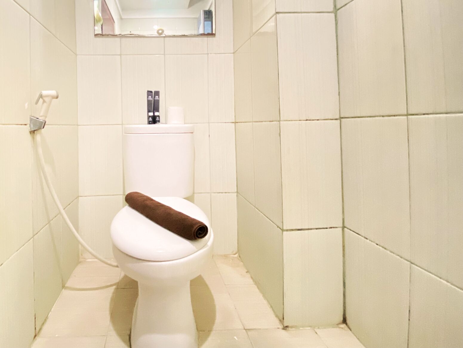 Sanitário pequeno e branco com uma toalha castanha em cima, um suporte branco para papel higiénico e um cano visível à esquerda.