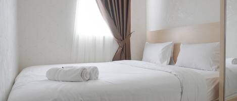 Cama bem arrumada com lençóis brancos e uma toalha enrolada, cabeceira de madeira e janela com cortinas leves.