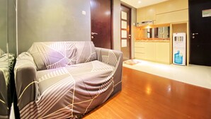 Sala de estar com sofá, área de cozinha com refrigerador e piso de madeira.