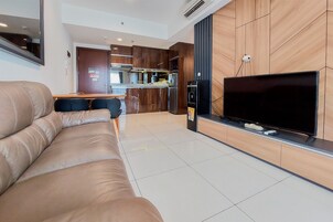 Sala de estar moderna com sofá de couro, televisão de tela plana e área de cozinha ao fundo.