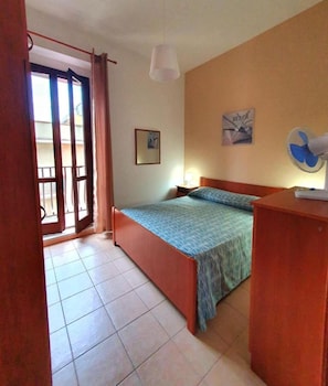 Quarto com cama, mesa de cabeceira, ventilador e janela com cortinas.