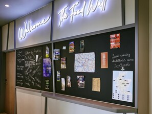 Paredes com um letreiro de neon 'Welcome', uma lousa com anotações à mão e diversos materiais impressos, incluindo mapas e folhetos.