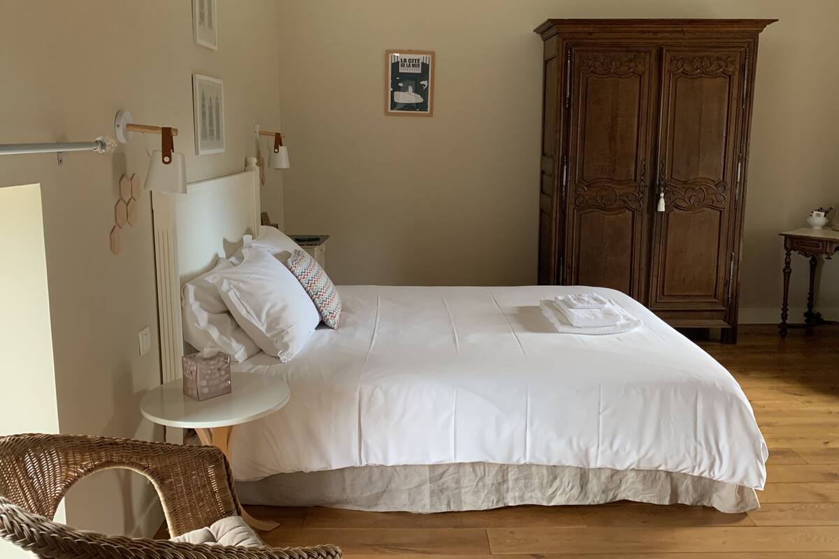 Quarto com uma cama grande, um guarda-roupa de madeira, uma mesinha redonda e uma cadeira de vime.