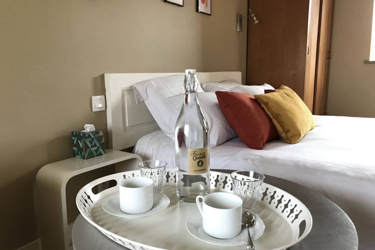 Cama bem arrumada com almofadas e mesa de cabeceira com bandeja contendo uma garrafa, xícaras e uma colher.