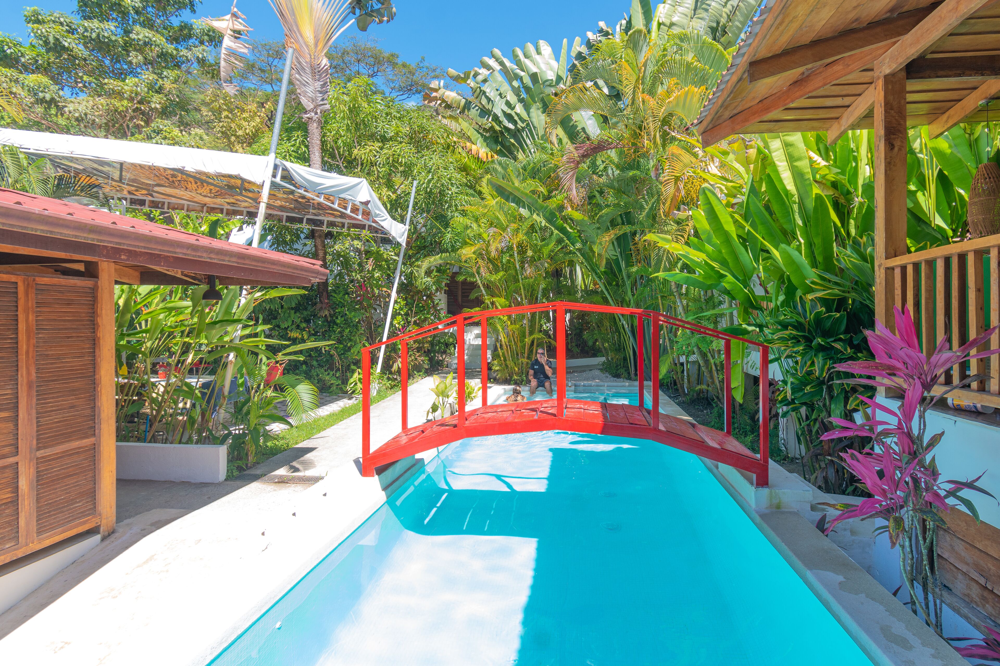 Uma piscina com uma ponte vermelha, rodeada por vegetação tropical exuberante e estruturas de madeira.