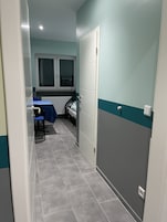 Um corredor estreito com uma cama, uma cadeira e uma janela.