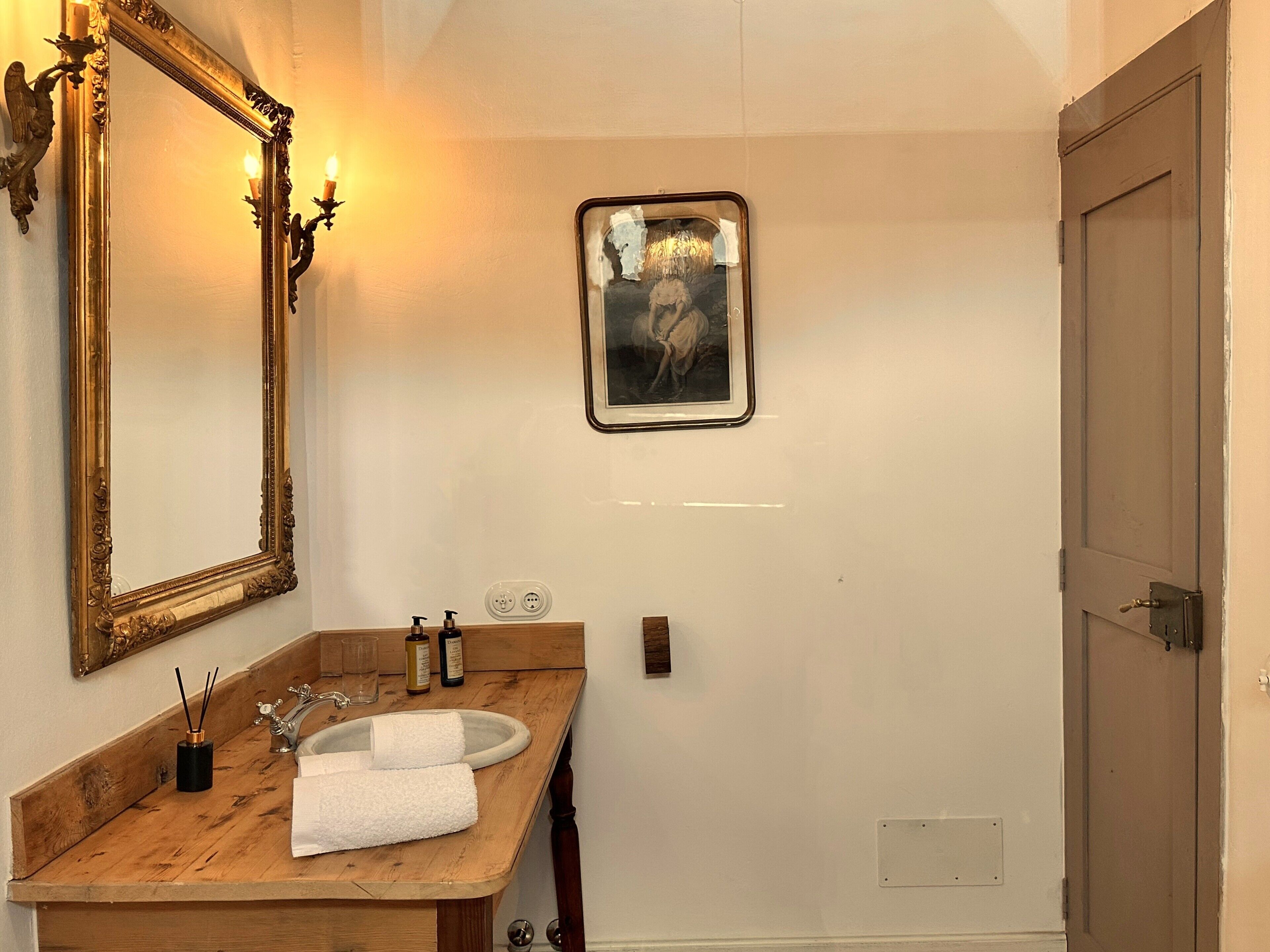 Casa de banho com toucador de madeira, um espelho grande e ornamentado, um quadro na parede e uma porta.
