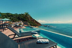Piscina infinita no terraço com espreguiçadeiras, vista para o oceano e montanhas ao fundo.