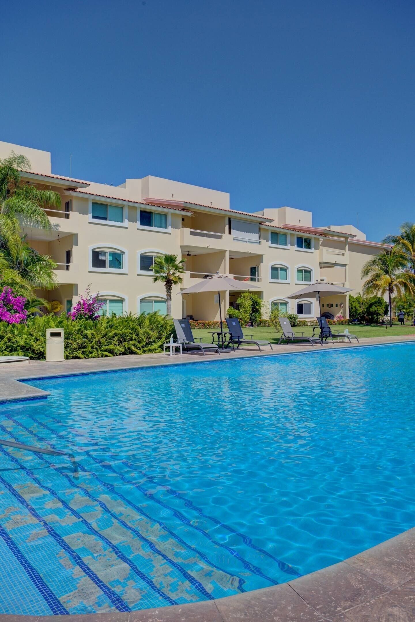 Piscina com espreguiçadeiras e um edifício ao fundo com várias varandas.